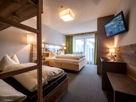 Quarto de hotel com uma cama beliche de madeira, uma cama de solteiro, uma escrivaninha e uma televisão presa na parede.