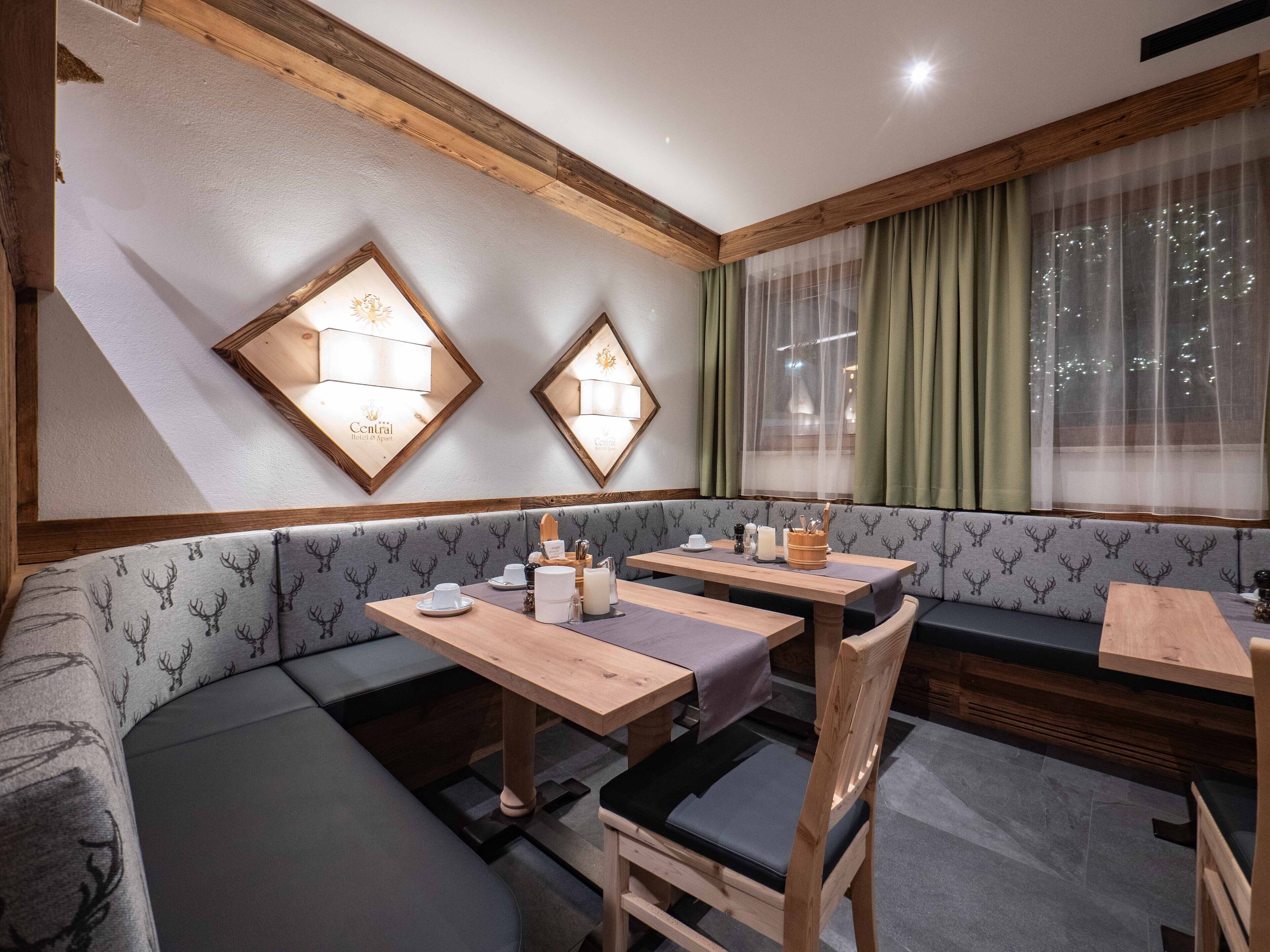 Um restaurante acolhedor com mesas de madeira e bancos estofados, arandelas nas paredes e uma grande janela com cortinas leves.