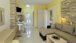 Uma sala de estar moderna com um sofá, uma mesa de centro e uma TV fixada na parede. Há uma área de cozinha com uma pia e bancada. O quarto possui uma parede de destaque em pedra e uma porta que dá para outro cômodo.