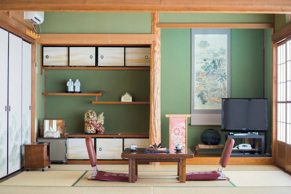 Um quarto japonês tradicional com tatames, uma mesa baixa, cadeiras, uma televisão e uma estante com objetos decorativos.