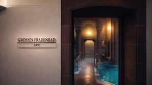 Entrada de hotel com uma placa indicando "GROSSES FRAUENBAD 34°C" e vista para uma área de piscina interna.