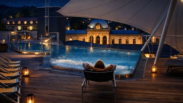 Uma pessoa relaxando em uma cadeira perto de uma piscina, com vista para um edifício com cúpula e arcos.