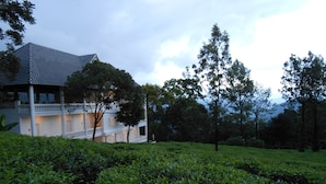 Um edifício de dois andares com varanda, rodeado por vegetação exuberante e árvores.