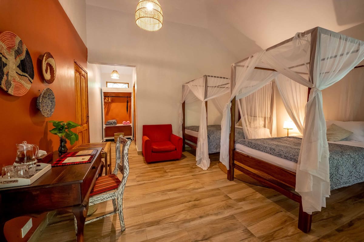 Um quarto com piso de madeira, uma escrivaninha com um vaso de flores, uma cadeira vermelha, uma cama com dossel e uma parede com cestos de palha.