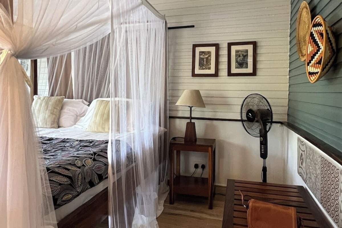 Quarto com cama de dossel, mesa de cabeceira com abajur, ventoinha na parede e duas imagens emolduradas.