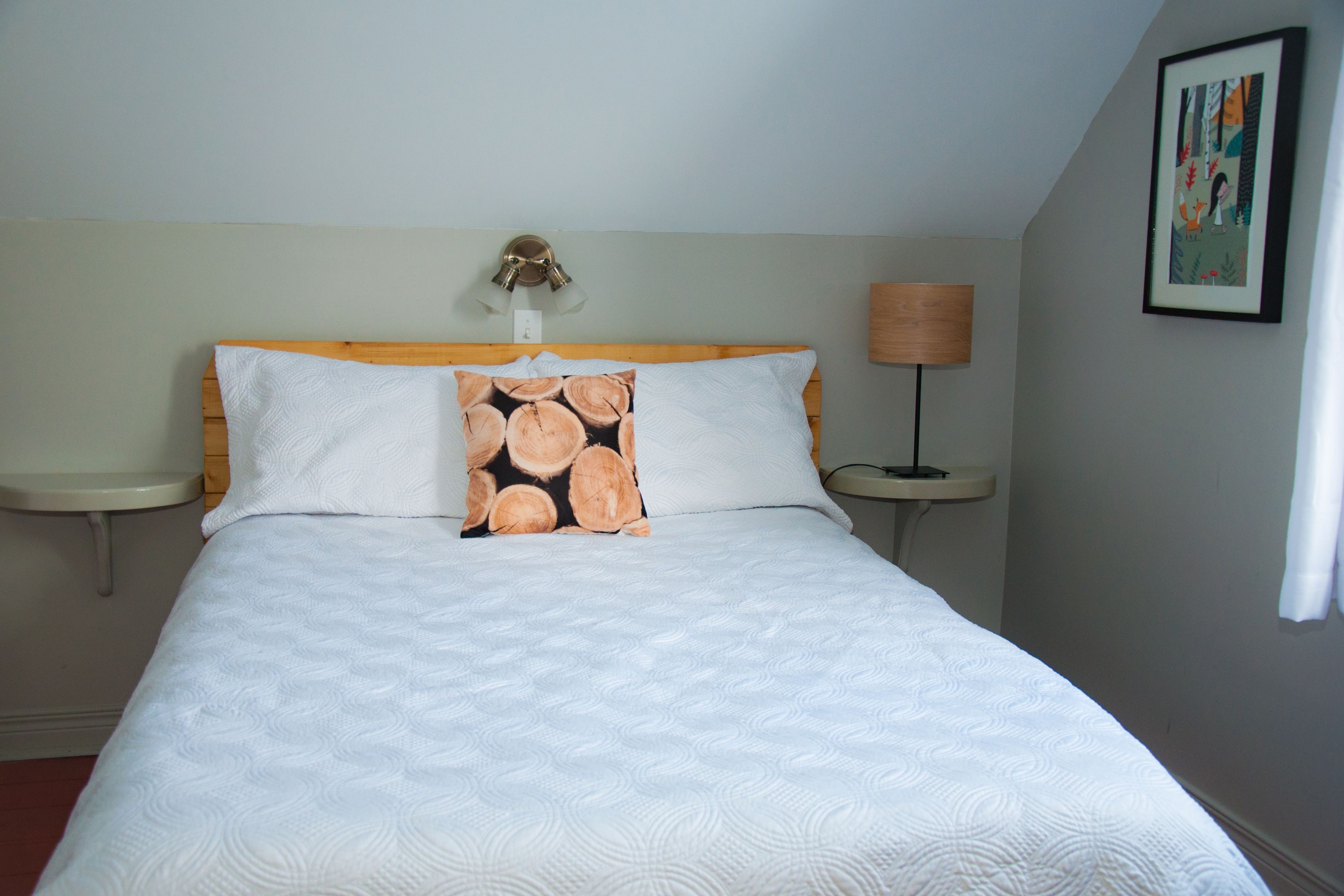 Cama bem arrumada com edredom branco e almofada estampada com padrão de madeira, luminária de cabeceira e quadro na parede.