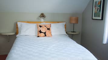 Cama bem arrumada com edredom branco e almofada estampada com padrão de madeira, luminária de cabeceira e quadro na parede.