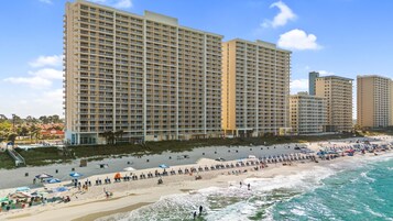 Um hotel à beira-mar com vários edifícios altos, uma praia arenosa com guarda-s óis e espreguiçadeiras, e vista para o oceano.