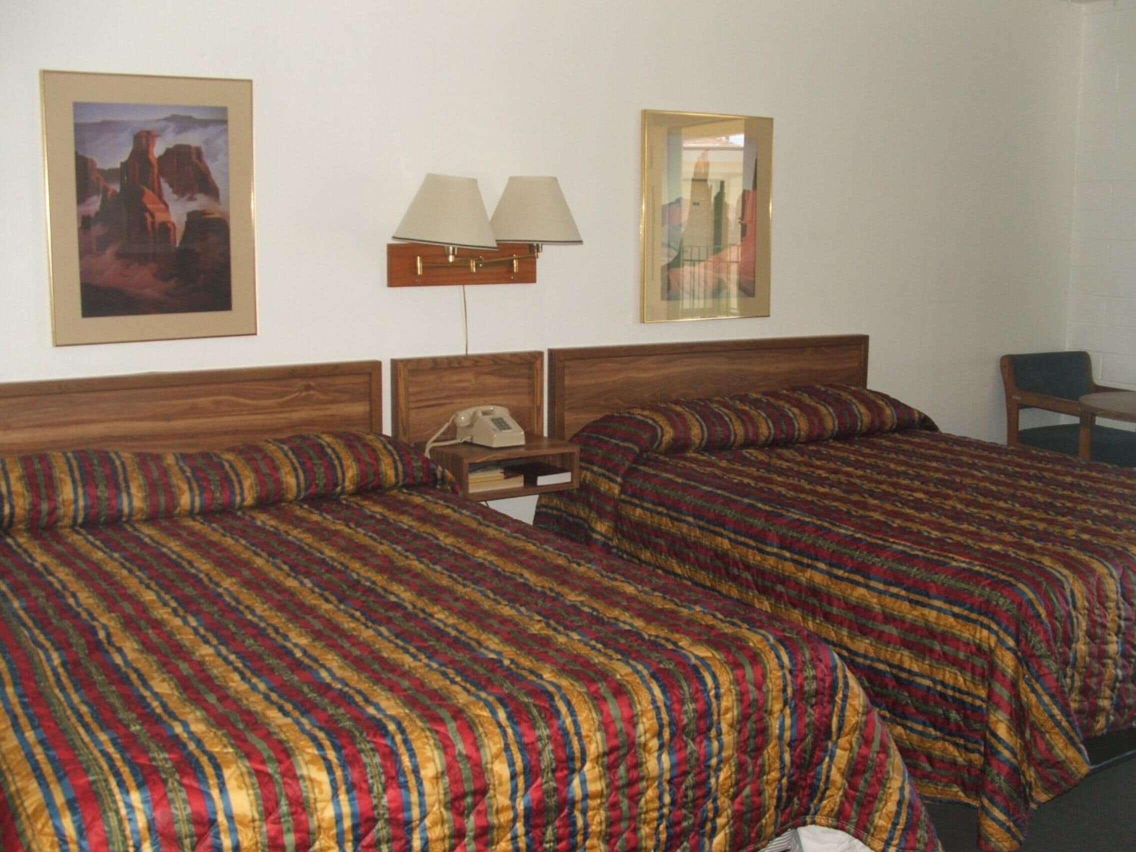 Quarto de hotel com duas camas, uma mesinha de cabeceira com abajur, um quadro na parede e uma cadeira.