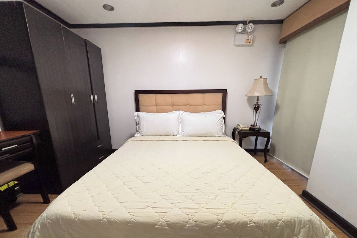 Uma cama bem arrumada com cabeceira acolchoada, uma mesinha de cabeceira com abajur e um guarda-roupa.