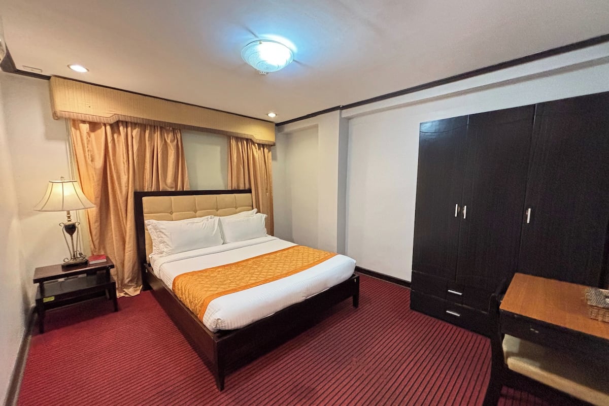Quarto de hotel com cama, mesa de cabeceira, luminária, guarda-roupa e escrivaninha.