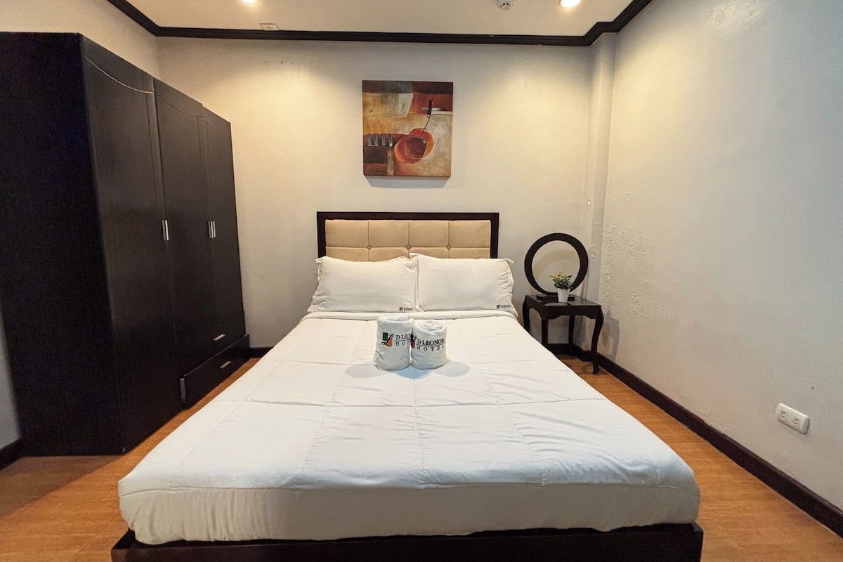 Cama bem arrumada com lençóis brancos, cabeceira e um quadro em cima. Um guarda-roupa escuro à esquerda e uma pequena mesa com um espelho à direita.