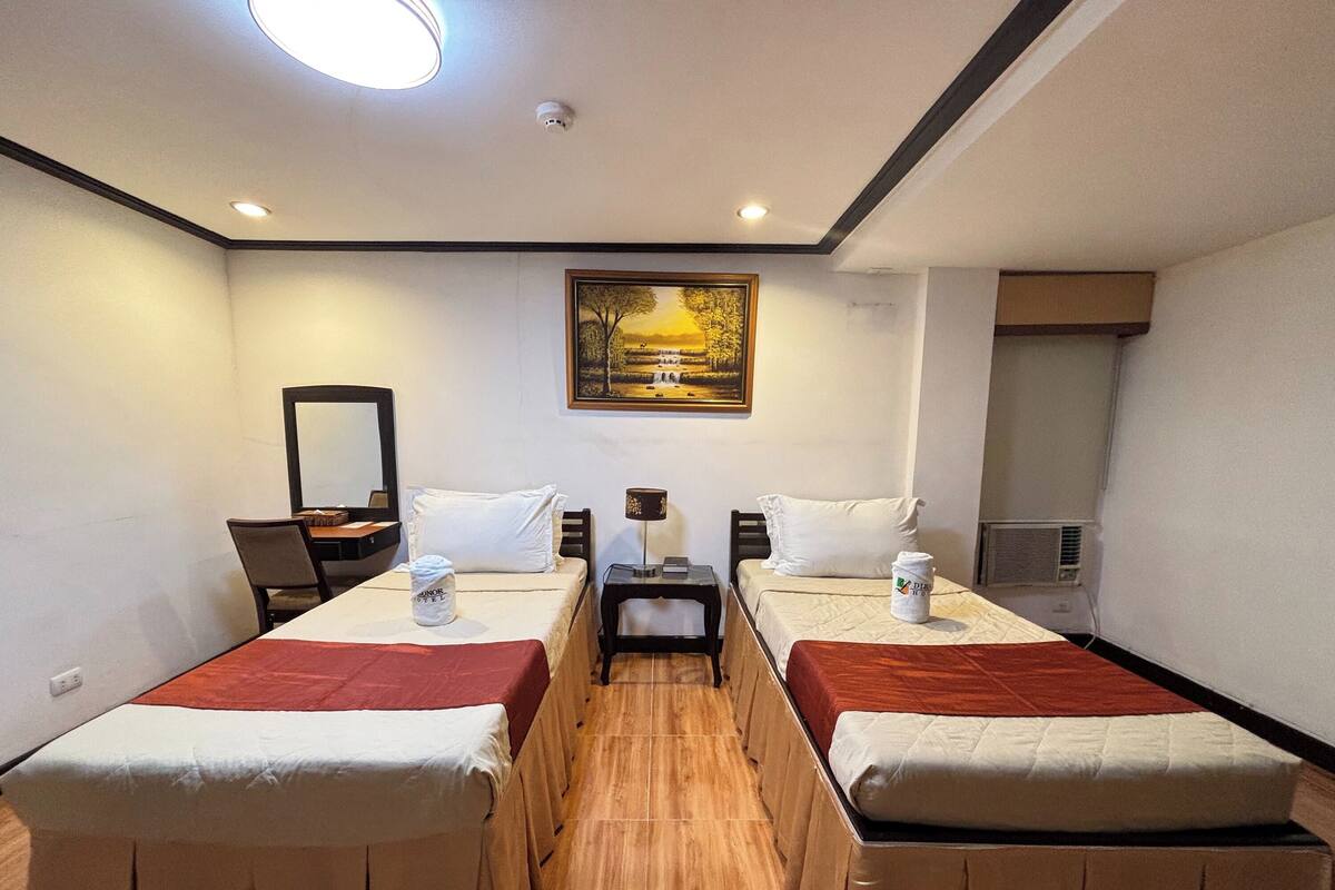 Quarto de hotel com duas camas de solteiro, uma escrivaninha, uma cadeira, um espelho e um quadro na parede.