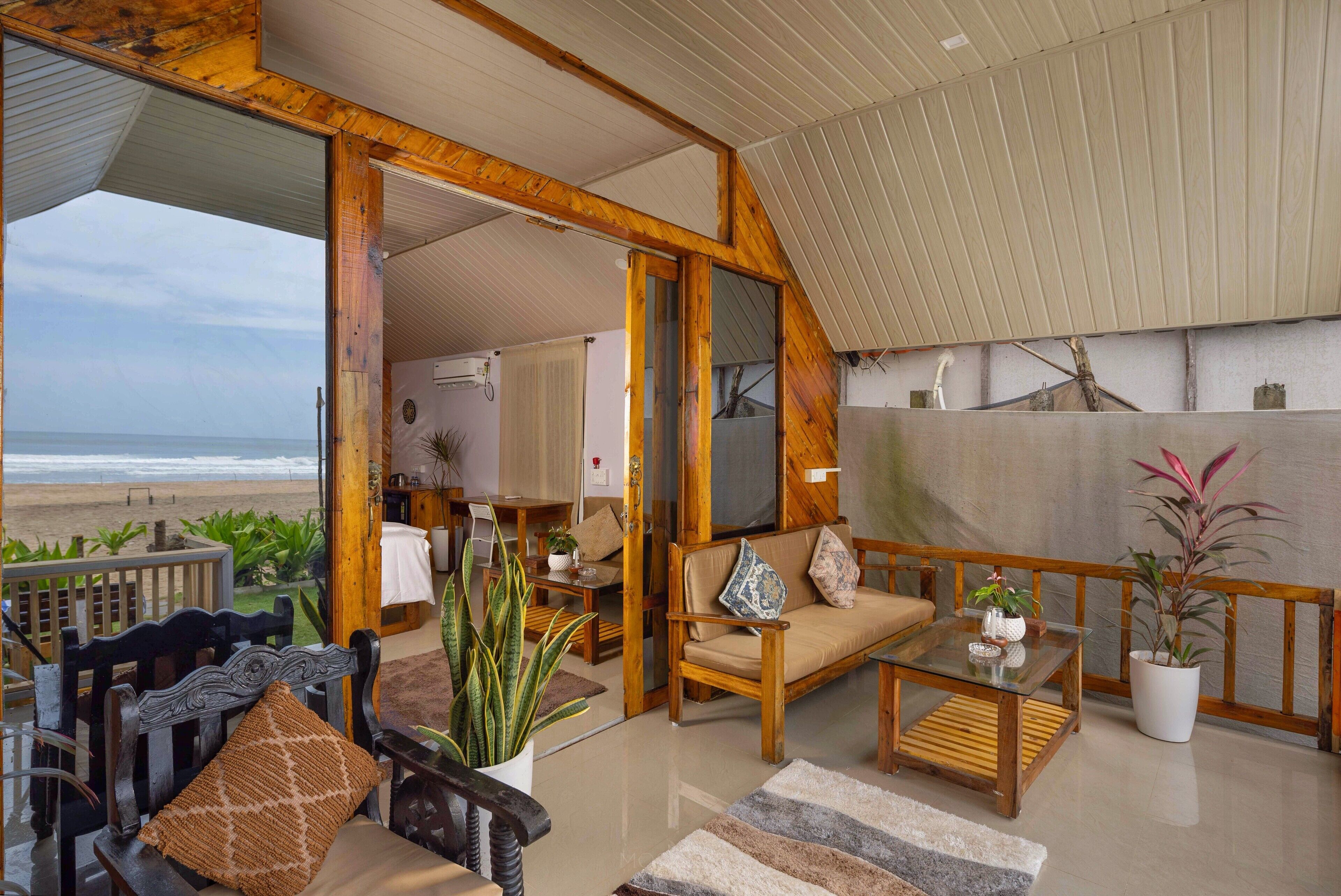 Ampla sala de estar com vista para a praia, móveis de madeira e plantas em vasos.
