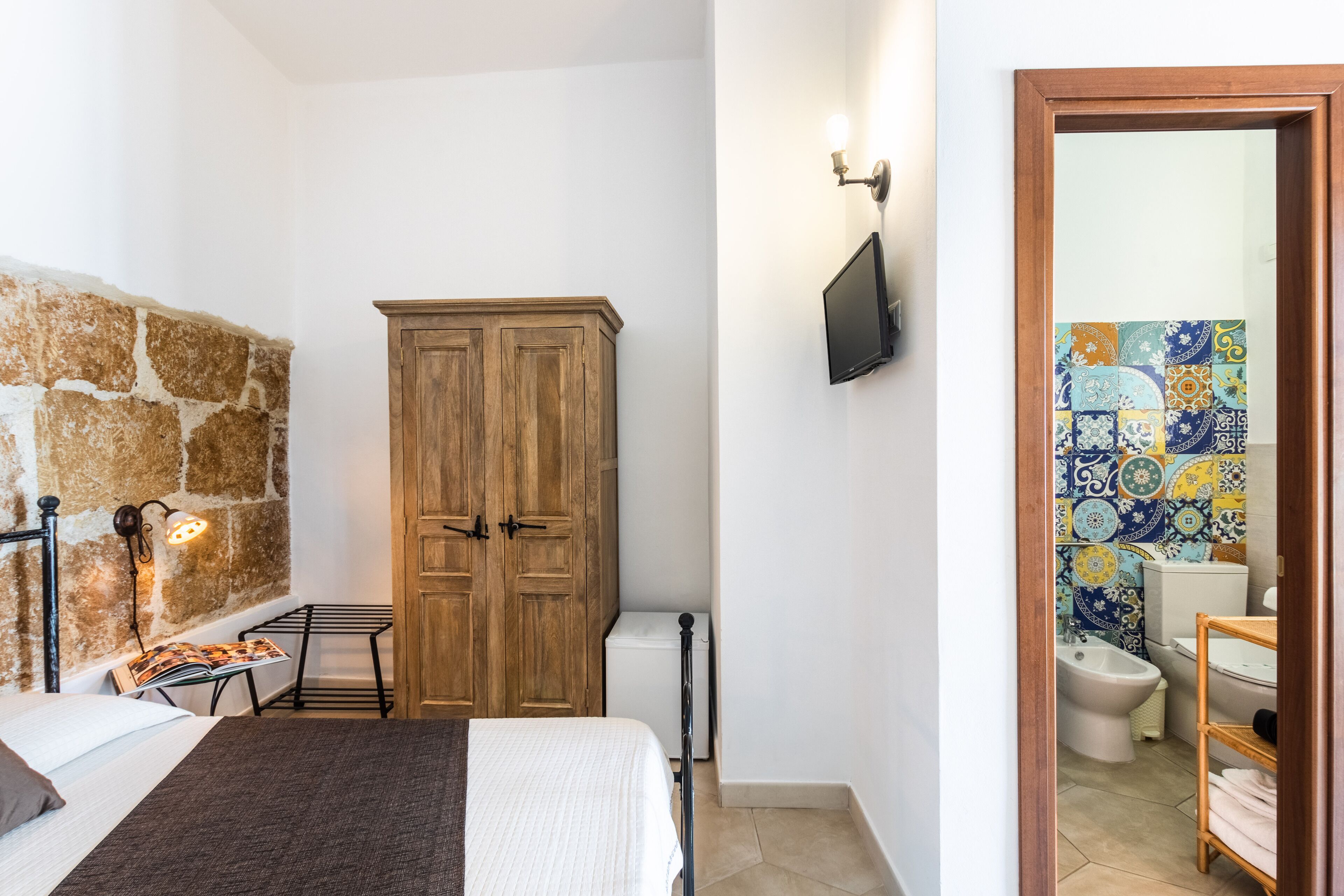 Quarto com cama, guarda-roupa de madeira, mesinha, luminária de leitura e televisão na parede.