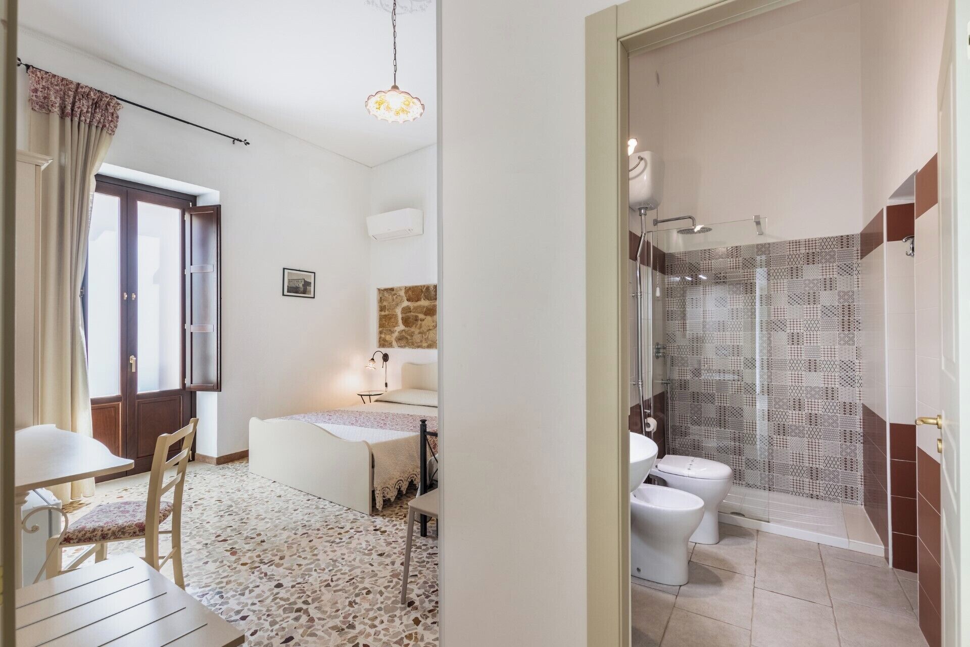 Quarto com cama, mesa, cadeira, luminária, janela com cortinas e banheiro com chuveiro, vaso sanitário e paredes azulejadas.