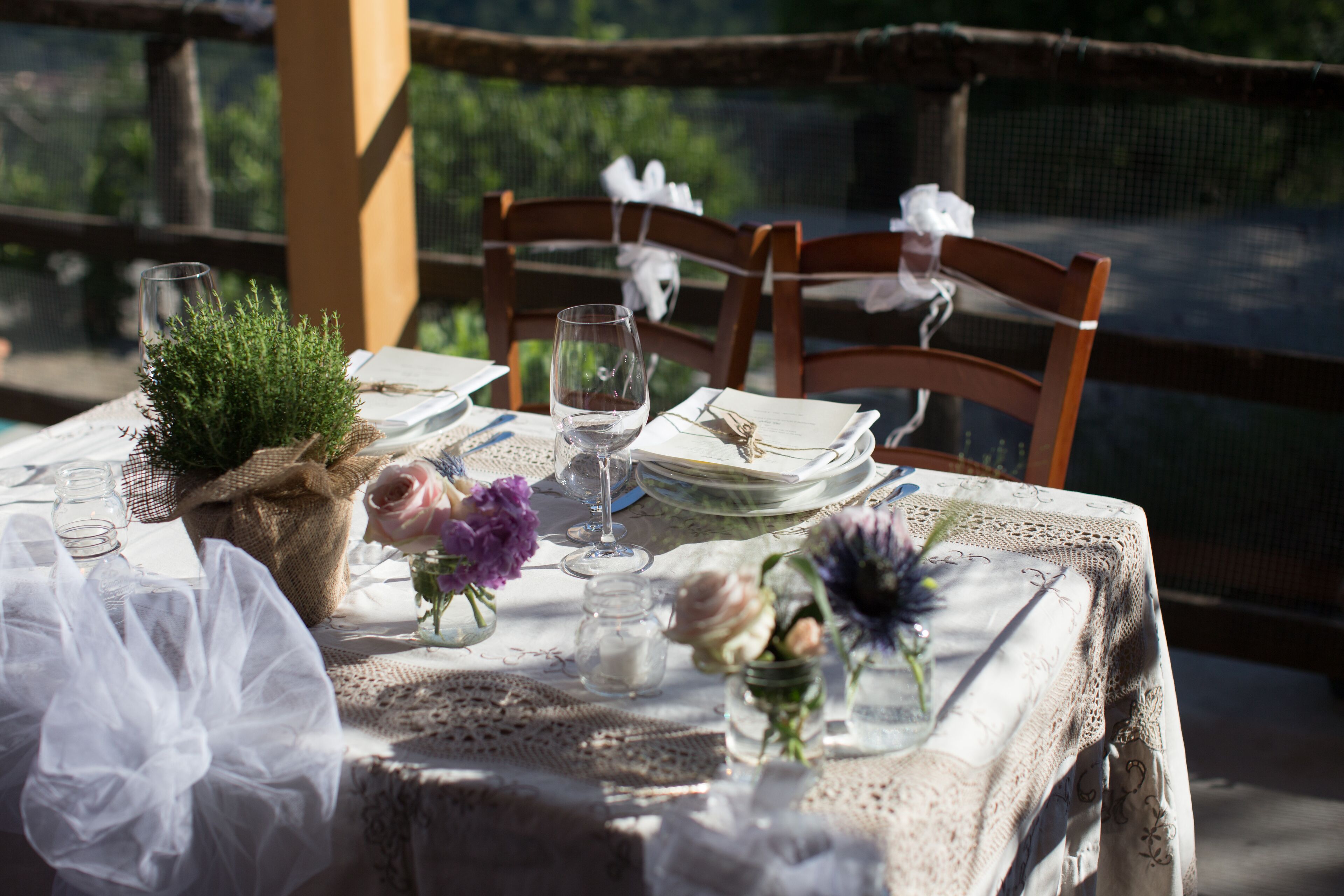 Mesa posta para refeição com toalha de renda, guardanapos brancos e centros de mesa florais.