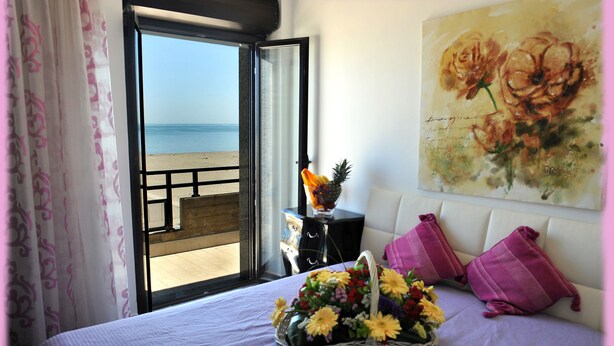 Um quarto com cama, janela vista para a praia, um quadro na parede e um cesto de flores.