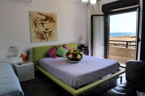 Quarto de hotel com cama, mesa de cabeceira, luminária e vista para a praia.