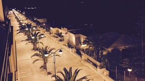 Vista noturna de um longo e bem iluminado calçadão, com palmeiras e postes de luz, que conduz a um edifício com telhado plano.