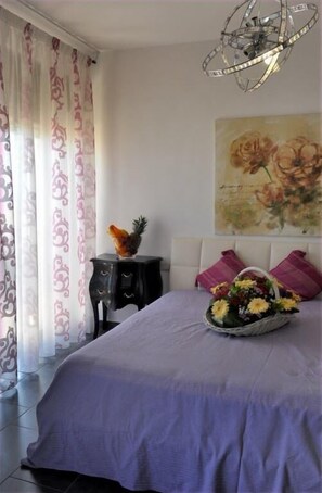 Quarto com colcha roxa, um lustre decorativo, uma pintura de flores e um vaso com flores num criado-mudo.