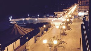Vista noturna de um calçadão à beira-mar, com postes de luz, palmeiras e edifícios ao longo da orla.