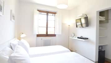 Quarto de hotel com duas camas, televisão fixada na parede, radiador e janela com molduras de madeira.