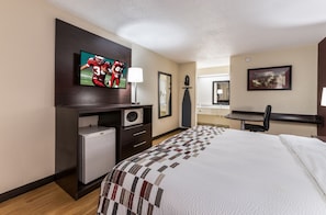 Quarto de hotel com cama, televisão, escrivaninha com micro-ondas, cadeira e pequeno frigorífico.