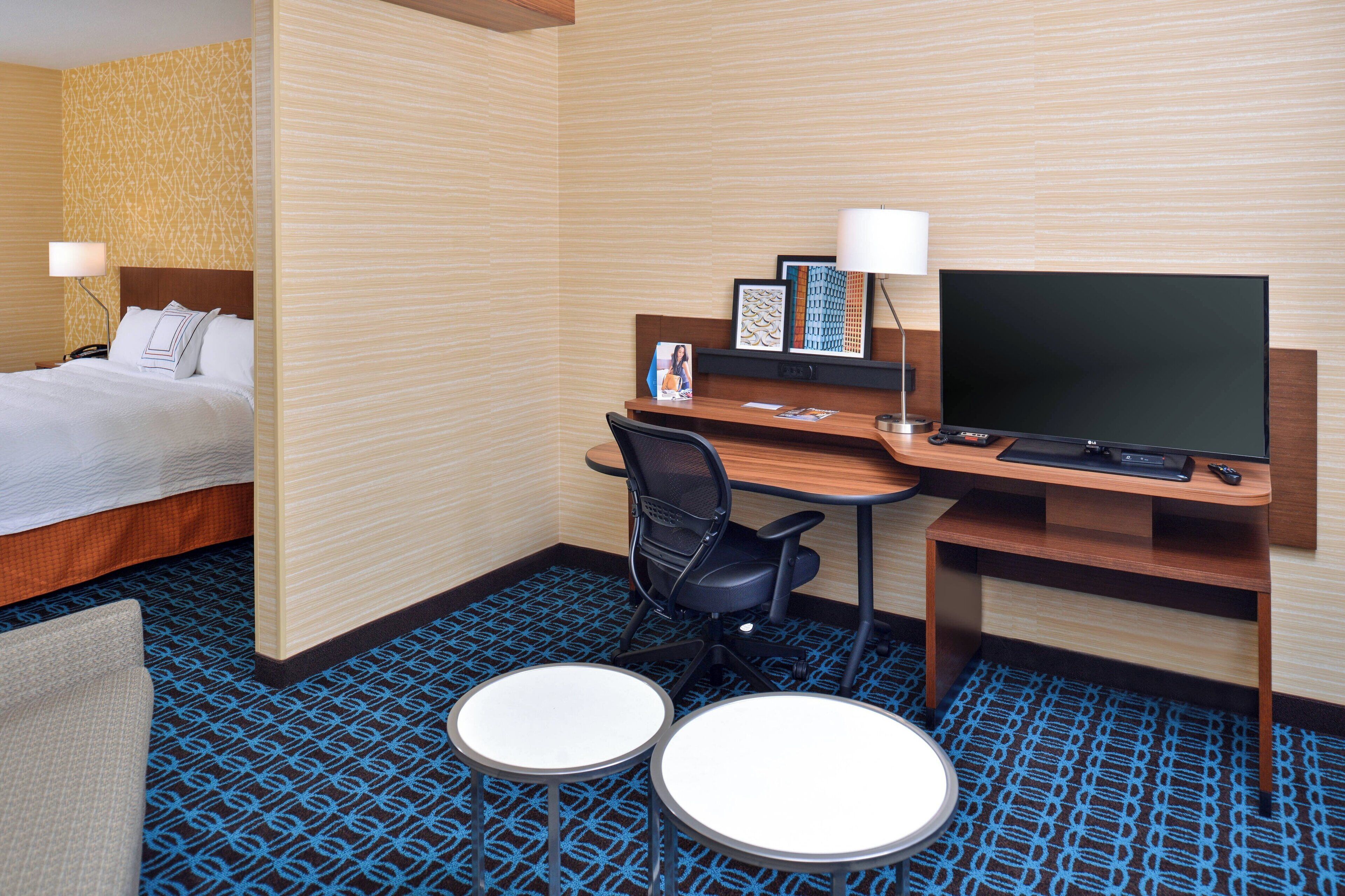 Quarto de hotel com uma cama, uma escrivaninha com computador, uma cadeira, duas mesinhas e uma televisão.