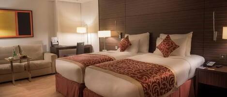 Quarto de hotel com duas camas, um sofá, uma escrivaninha e uma luminária.