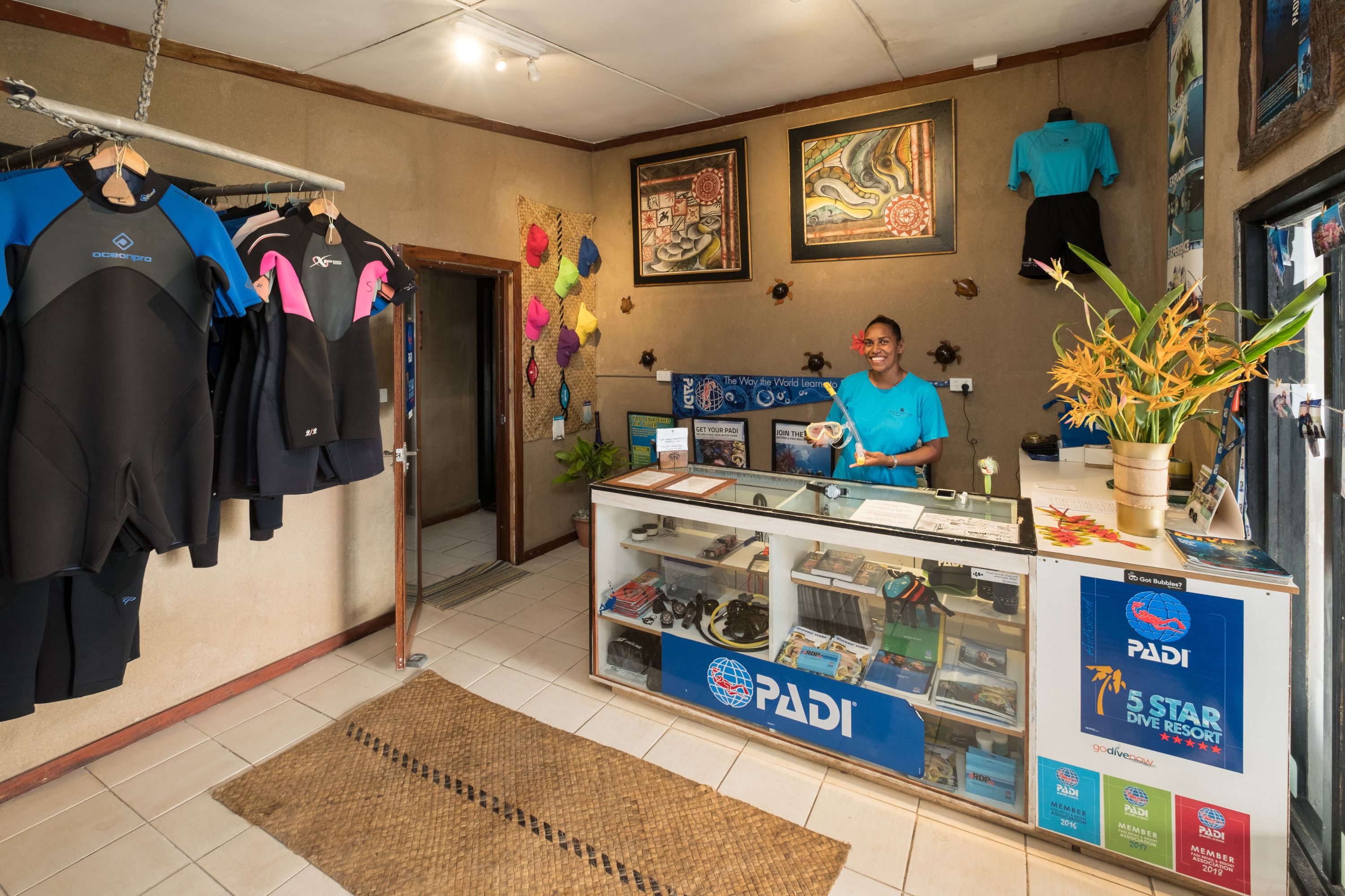 Interior de loja de mergulho, com fatos de neoprene, um balcão com material promocional da PADI e uma parede com obras de arte emolduradas.