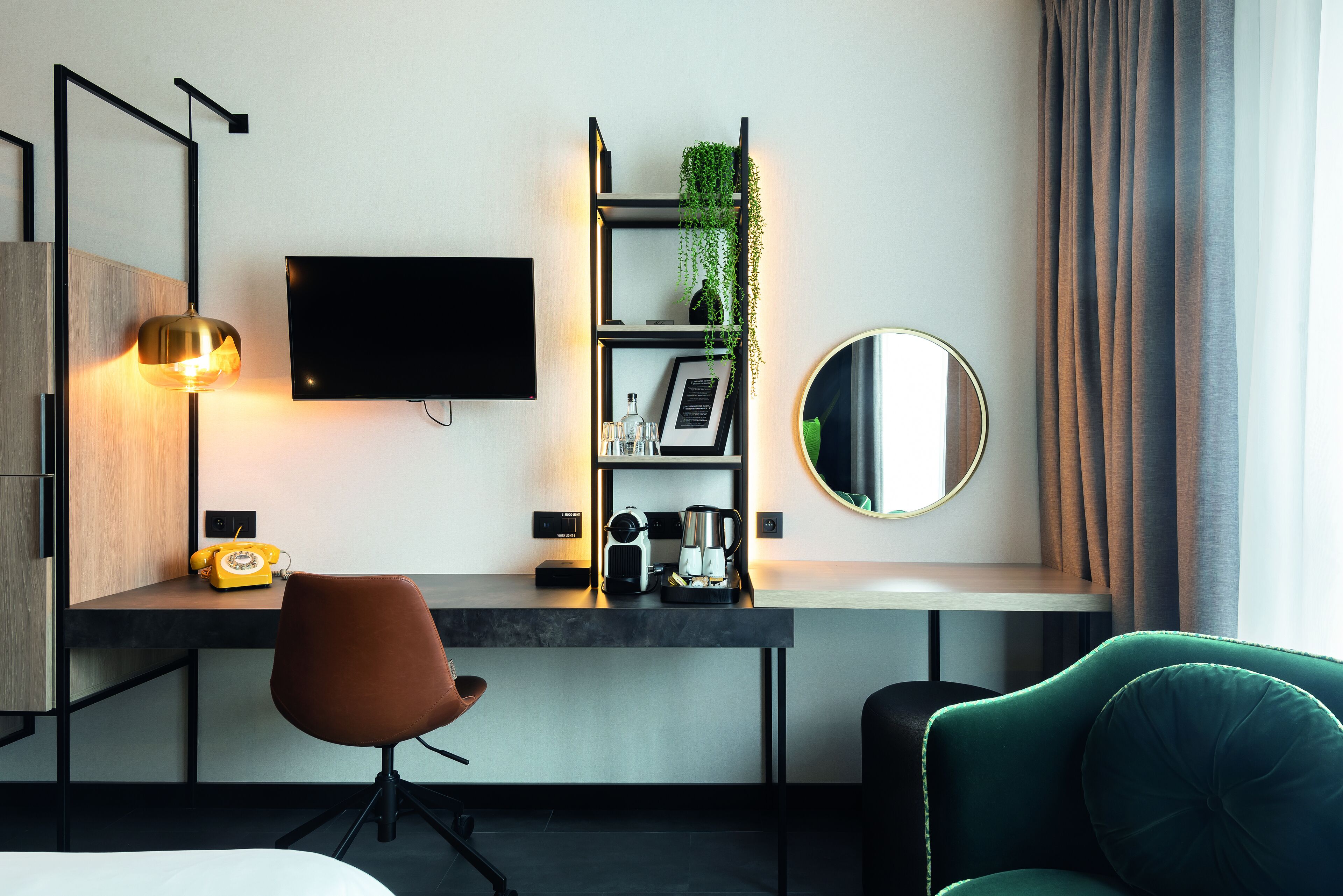 Quarto de hotel moderno com uma escrivaninha, televisão, cafeteira e uma planta em uma estante.