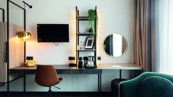 Quarto de hotel moderno com uma escrivaninha, televisão, cafeteira e uma planta em uma estante.