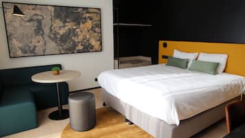 Quarto de hotel moderno com uma cama grande, uma mesinha redonda com uma planta, um sofá e uma lixeira.
