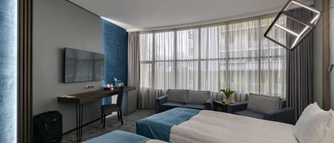 Quarto de hotel com duas camas, uma escrivaninha, televisão e uma janela grande com cortinas.