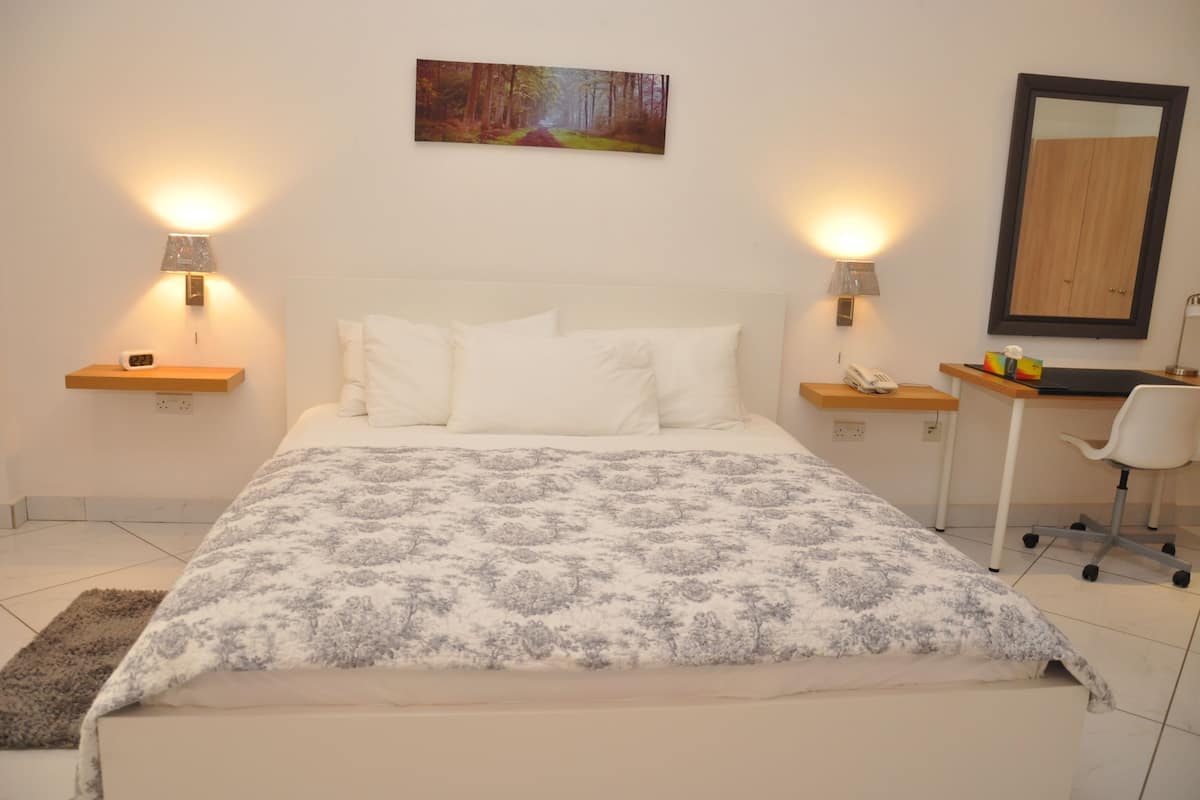 Quarto com cama, duas mesas de cabeceira, escrivaninha com cadeira, espelho e um quadro na parede.