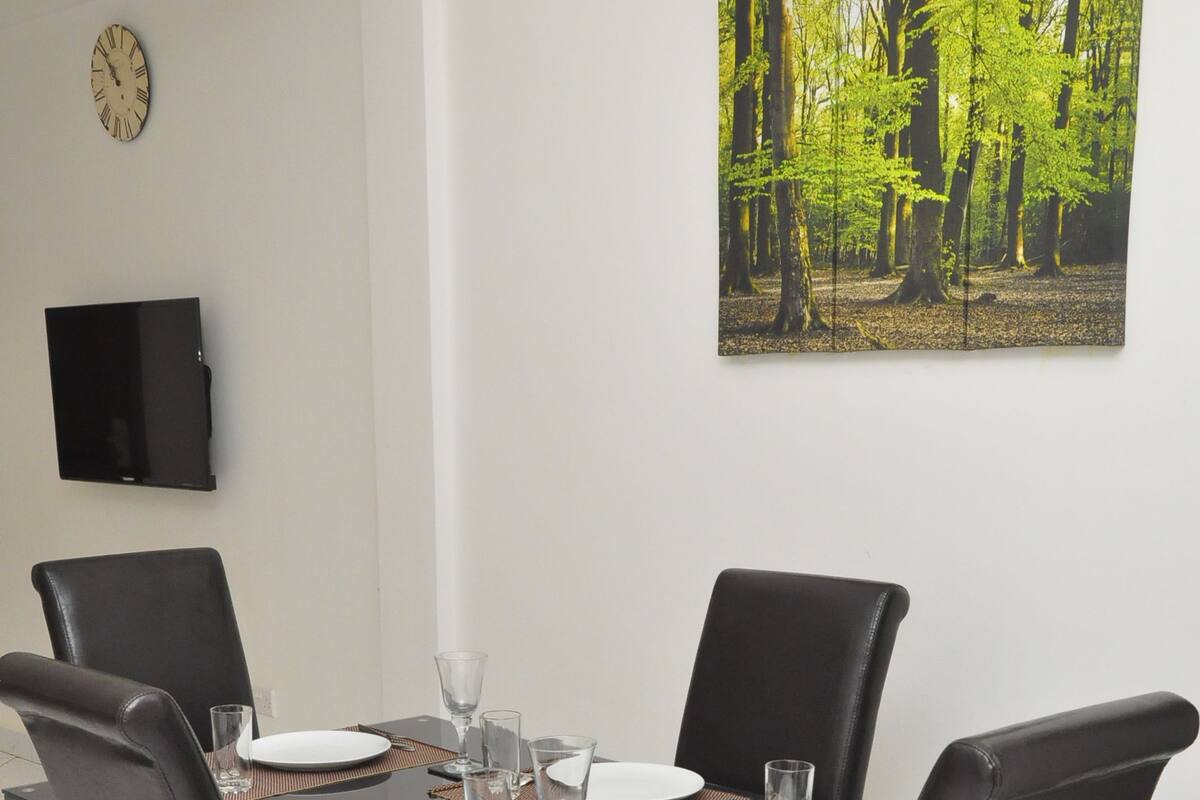 Uma mesa de jantar com cadeiras pretas, pratos brancos e copos, sobre um chão de azulejos, com um quadro de uma floresta na parede.