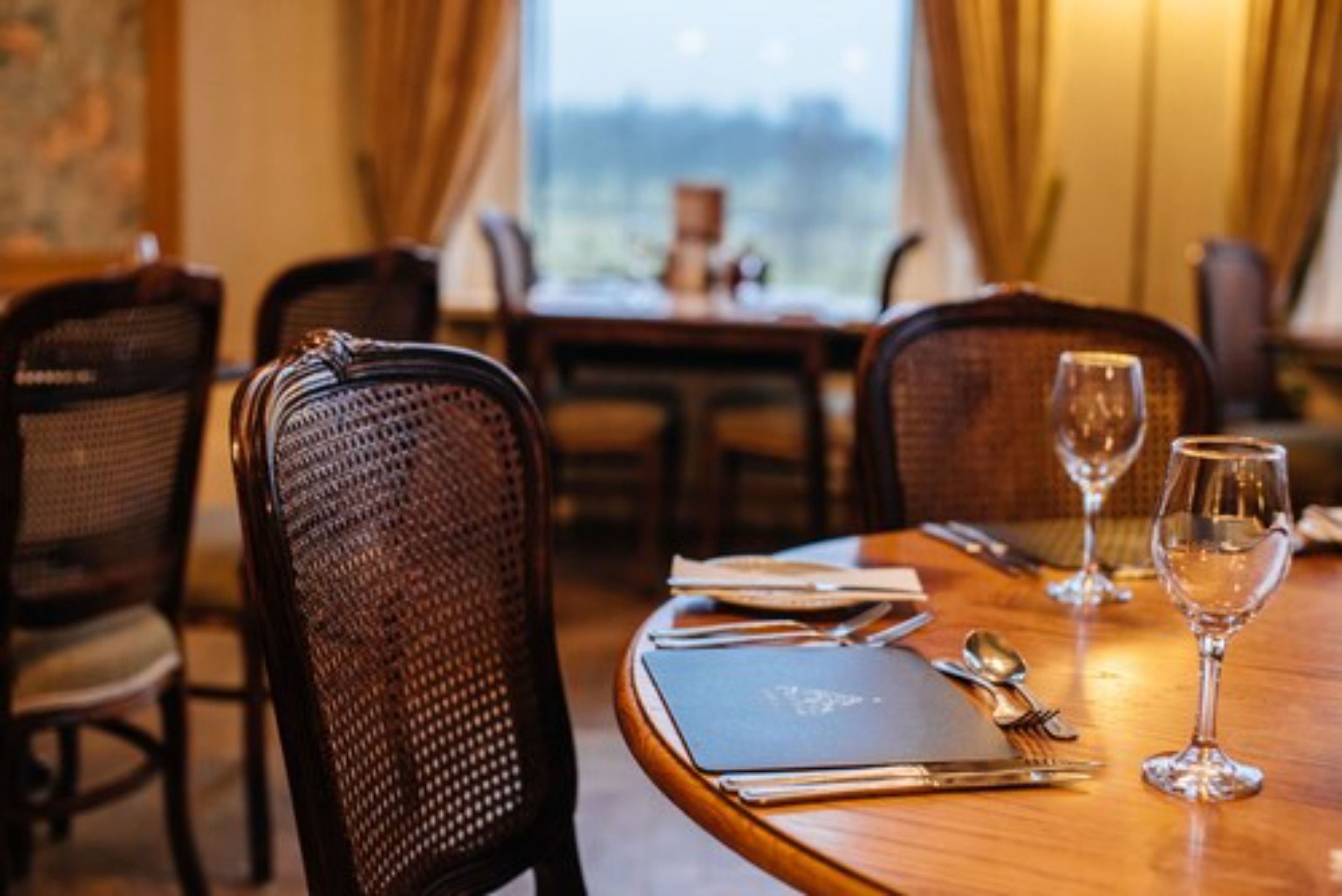 Mesa de jantar posta com menu, talheres e copos de vinho, com cadeiras de vime ao redor.