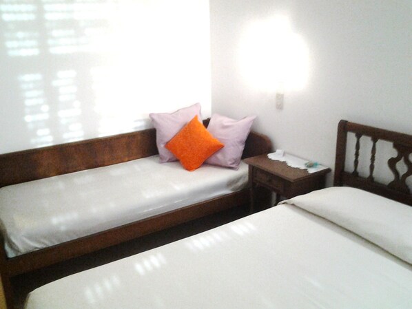 Quarto com duas camas, cada uma com roupa de cama branca e cabeceira de madeira. Há uma mesa de cabeceira com abajur entre as camas.