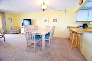 Uma sala de jantar com mesa e cadeiras de madeira, uma televisão e uma cozinha com cafeteira.