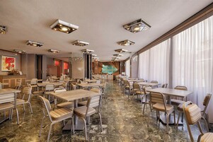 Uma sala de jantar ampla, com várias mesas e cadeiras, piso de mármore e iluminação no teto.