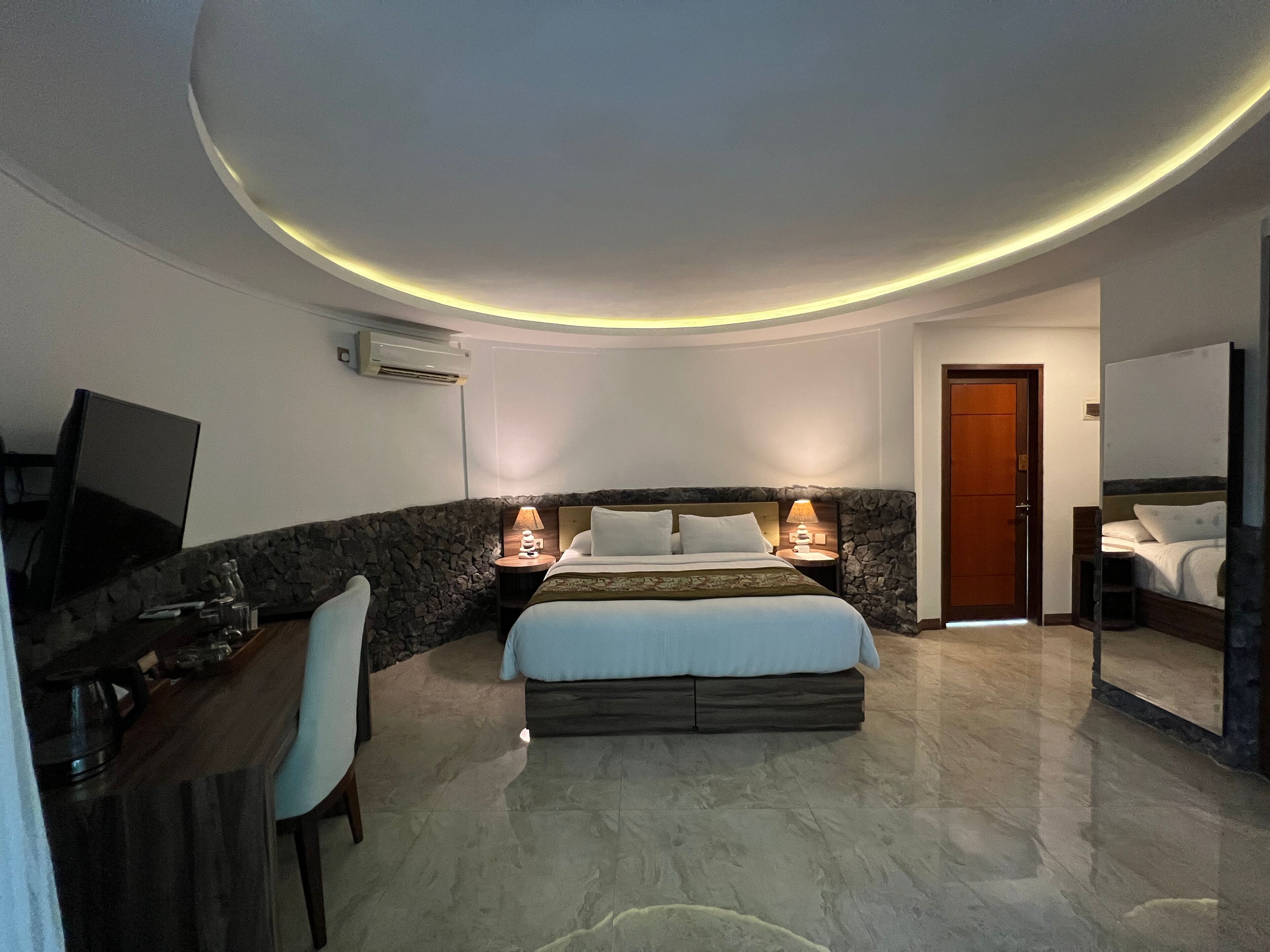 Quarto de hotel moderno com teto curvo, uma cama grande com duas mesas de cabeceira, televisão de tela plana e uma escrivaninha com cadeira.