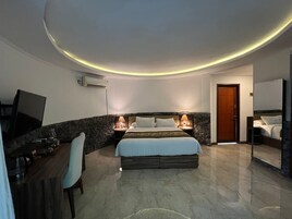 Quarto de hotel moderno com teto curvo, uma cama grande com duas mesas de cabeceira, televisão de tela plana e uma escrivaninha com cadeira.