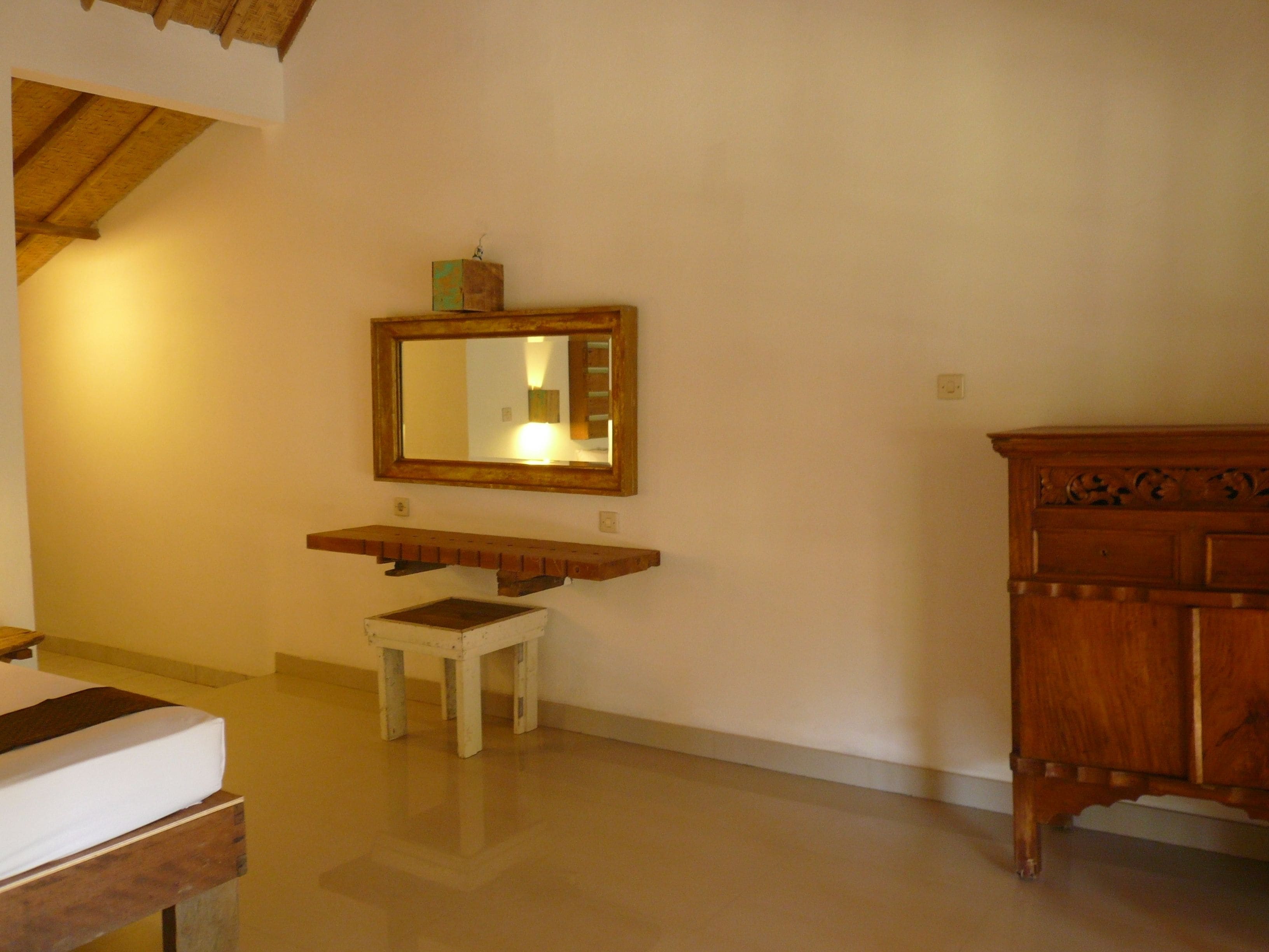 Quarto com penteadeira de madeira, um banquinho pequeno e um espelho na parede.