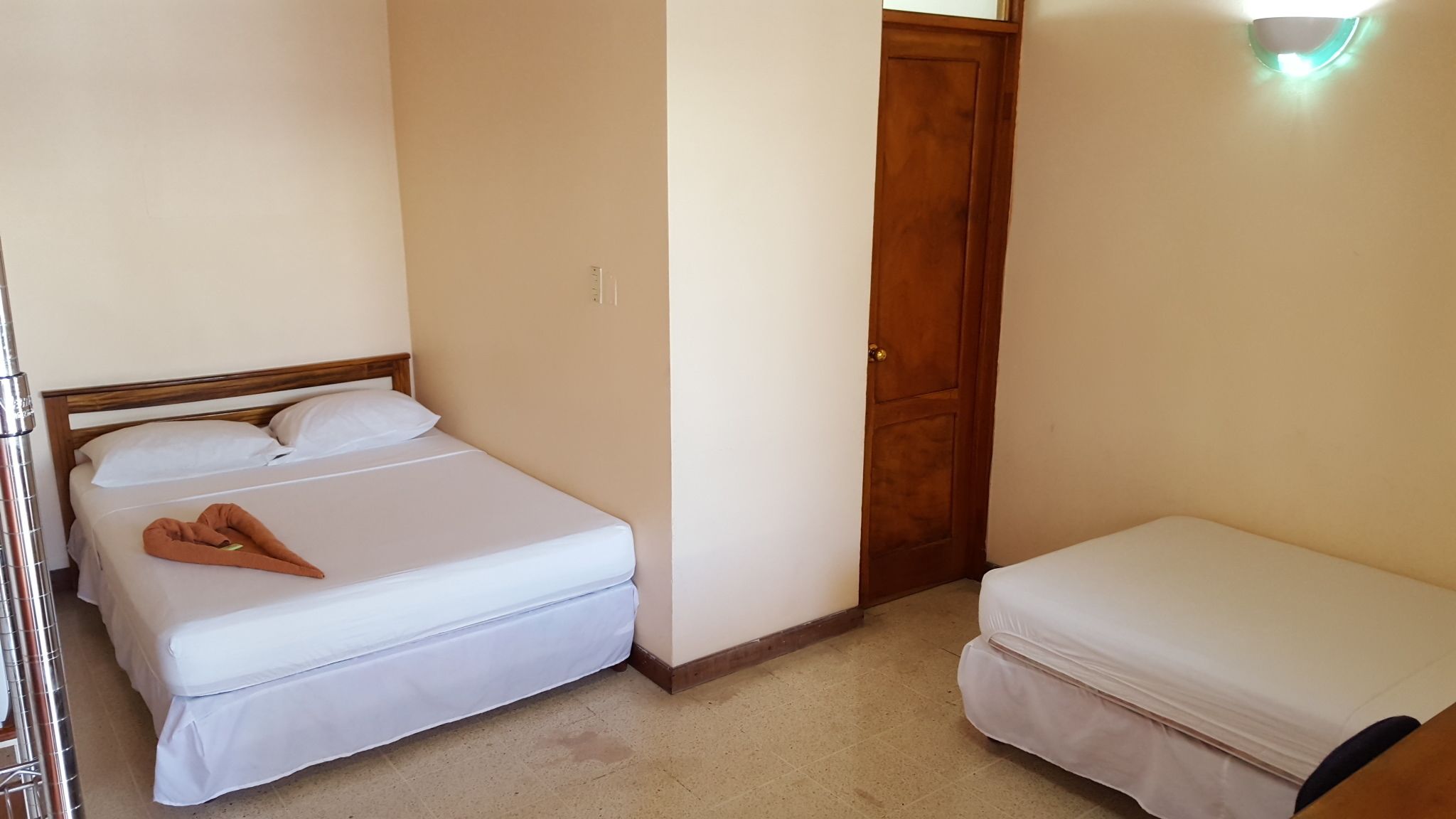 Quarto pequeno e limpo, com uma cama de solteiro, uma porta de madeira e uma pequena mesa.