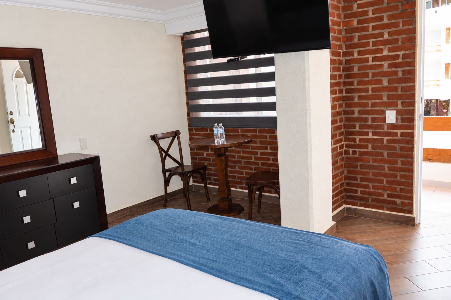 Quarto de hotel com cama, penteadeira, televisão presa à parede e parede de destaque em tijolo.