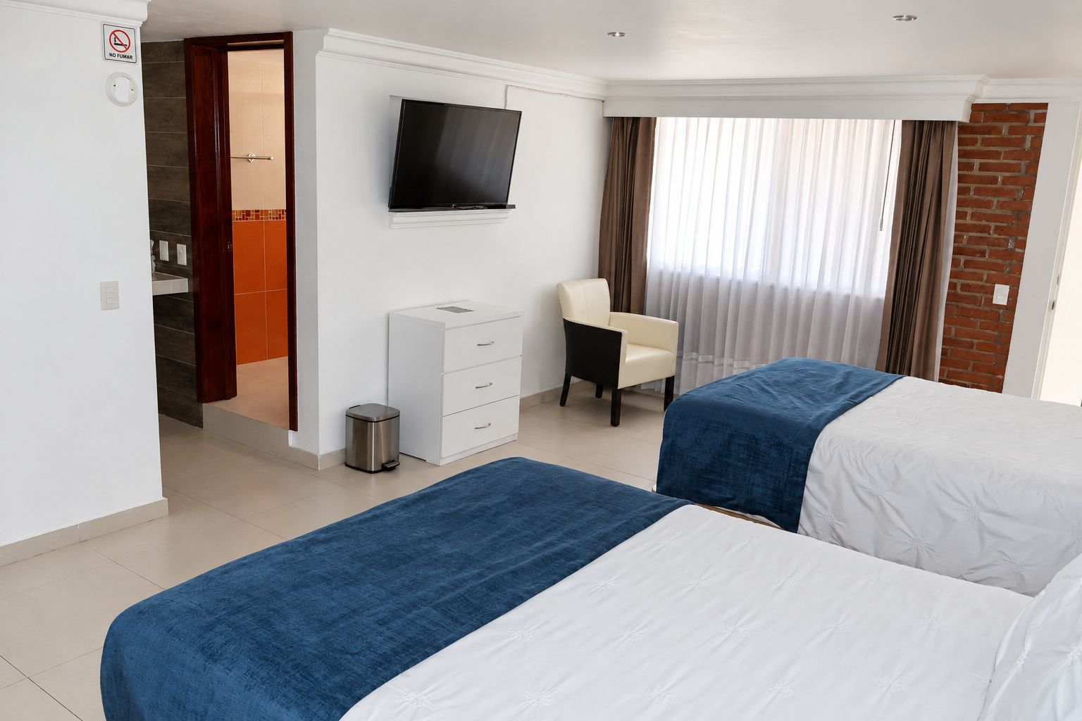 Quarto de hotel com duas camas, televisão, escrivaninha, cadeira e janela com cortinas.