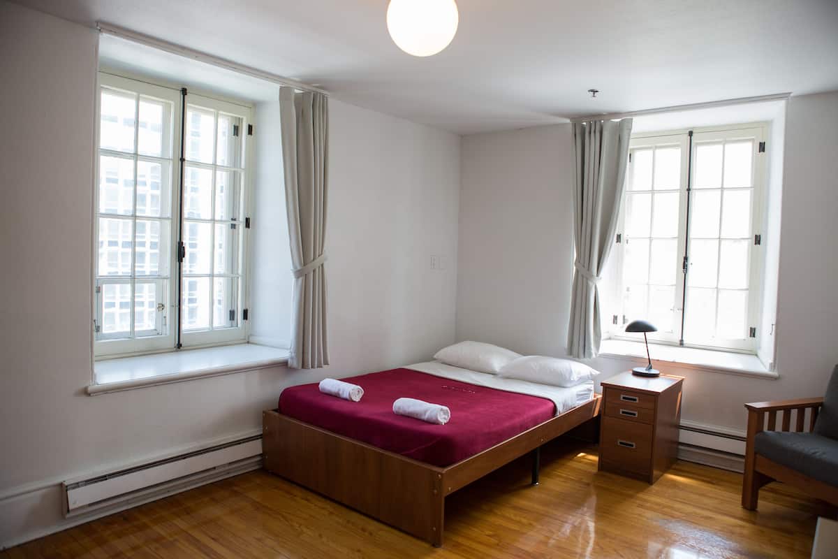 Quarto com cama de madeira, mesa de cabeceira, cadeira e duas janelas com cortinas.