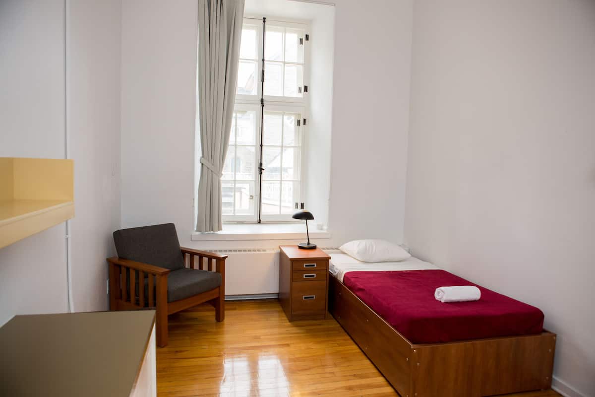 Quarto pequeno e bem iluminado com piso de madeira, uma cama de solteiro com cobertor vermelho, uma cadeira, uma mesinha com luminária e uma janela com cortinas.
