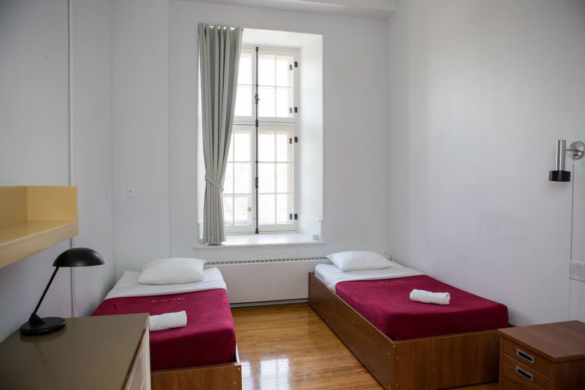 Quarto pequeno e limpo com duas camas de solteiro, uma janela com cortinas, uma escrivaninha com abajur e um radiador.